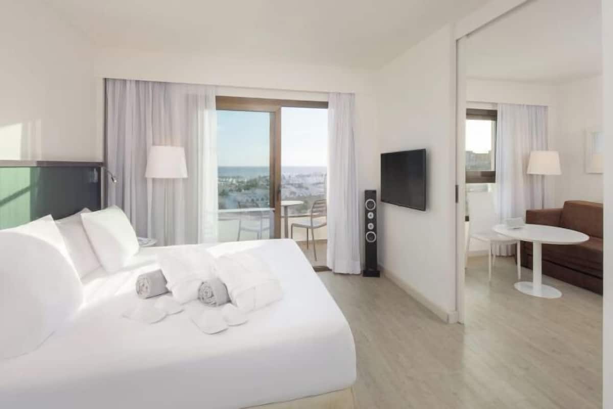 Quarto de hotel moderno com uma cama grande, televisão, varanda com vista e uma pequena área de estar.