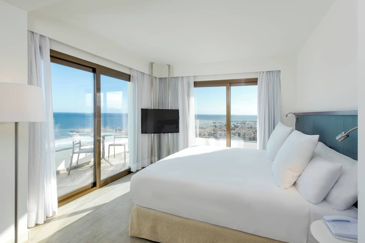 Quarto de hotel com uma cama grande, televisão de tela plana, escrivaninha com luminária e vista para o oceano.