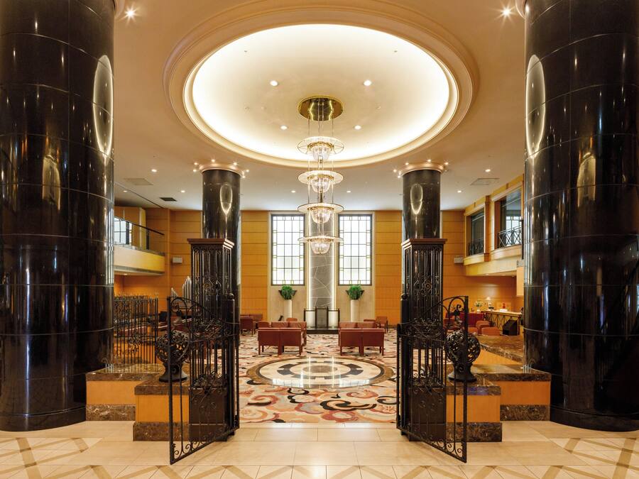 Um hall amplo com um lustre central, colunas pretas imponentes e um piso com padrão.