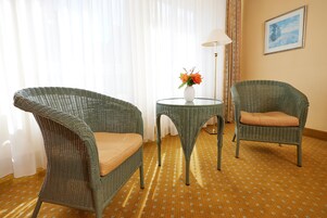 Quarto com duas cadeiras de vime, uma mesa de vidro pequena com um vaso de flores e um abajur.