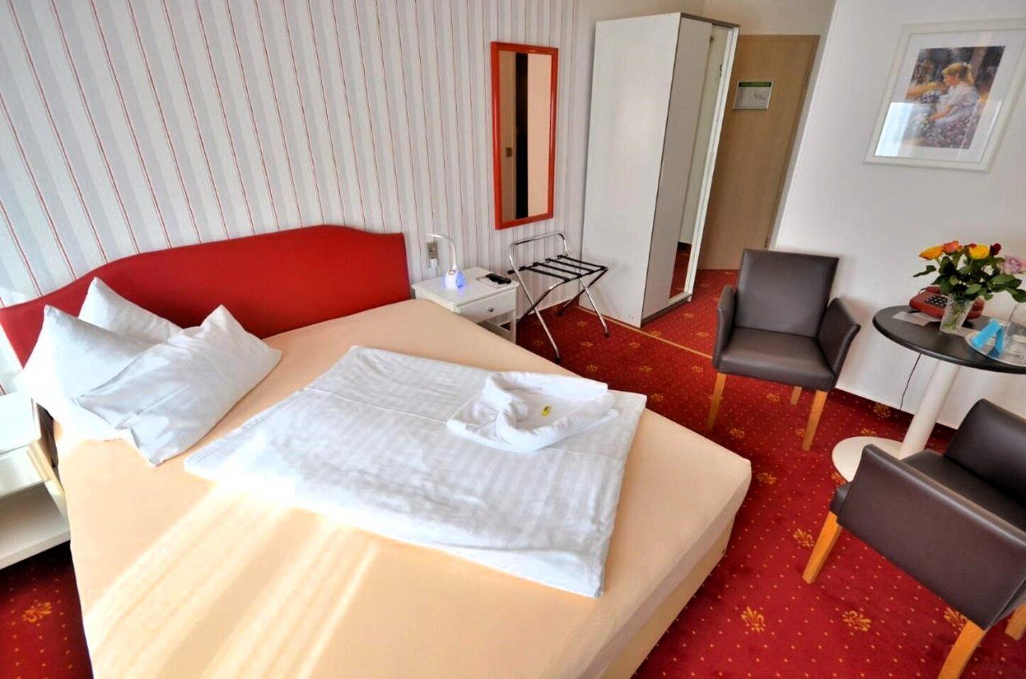 Quarto de hotel com cama, escrivaninha, cadeira e pequena mesa com um vaso de flores.