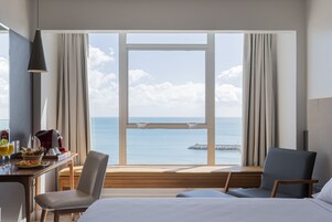 Quarto de hotel com uma janela ampla que oferece vista para o mar e um cais. Há uma cama, duas cadeiras, uma mesa com uma cafeteira e um tabuleiro de pequeno-almoço.