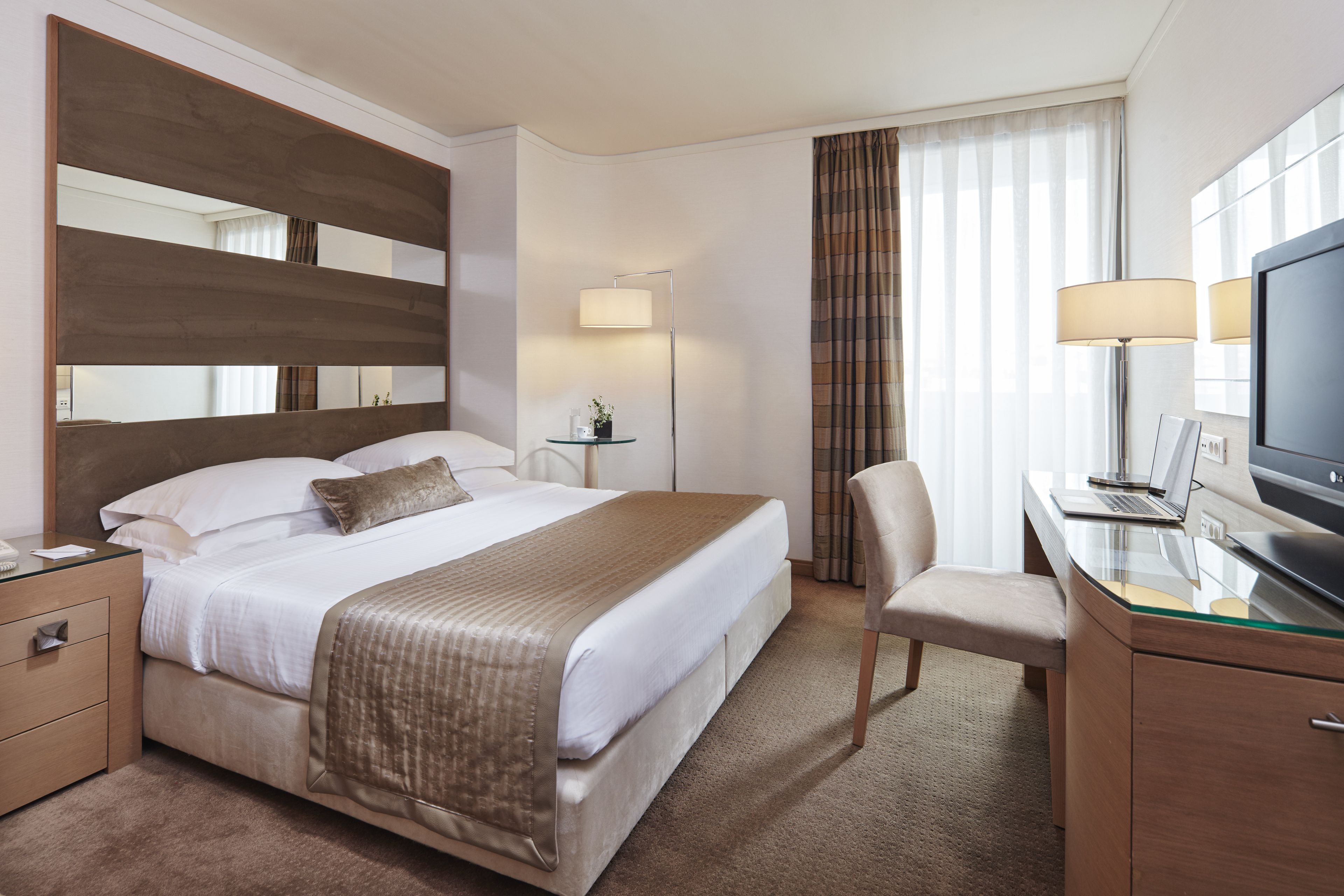 Quarto de hotel com uma cama grande, uma escrivaninha com computador, televisão e abajur.