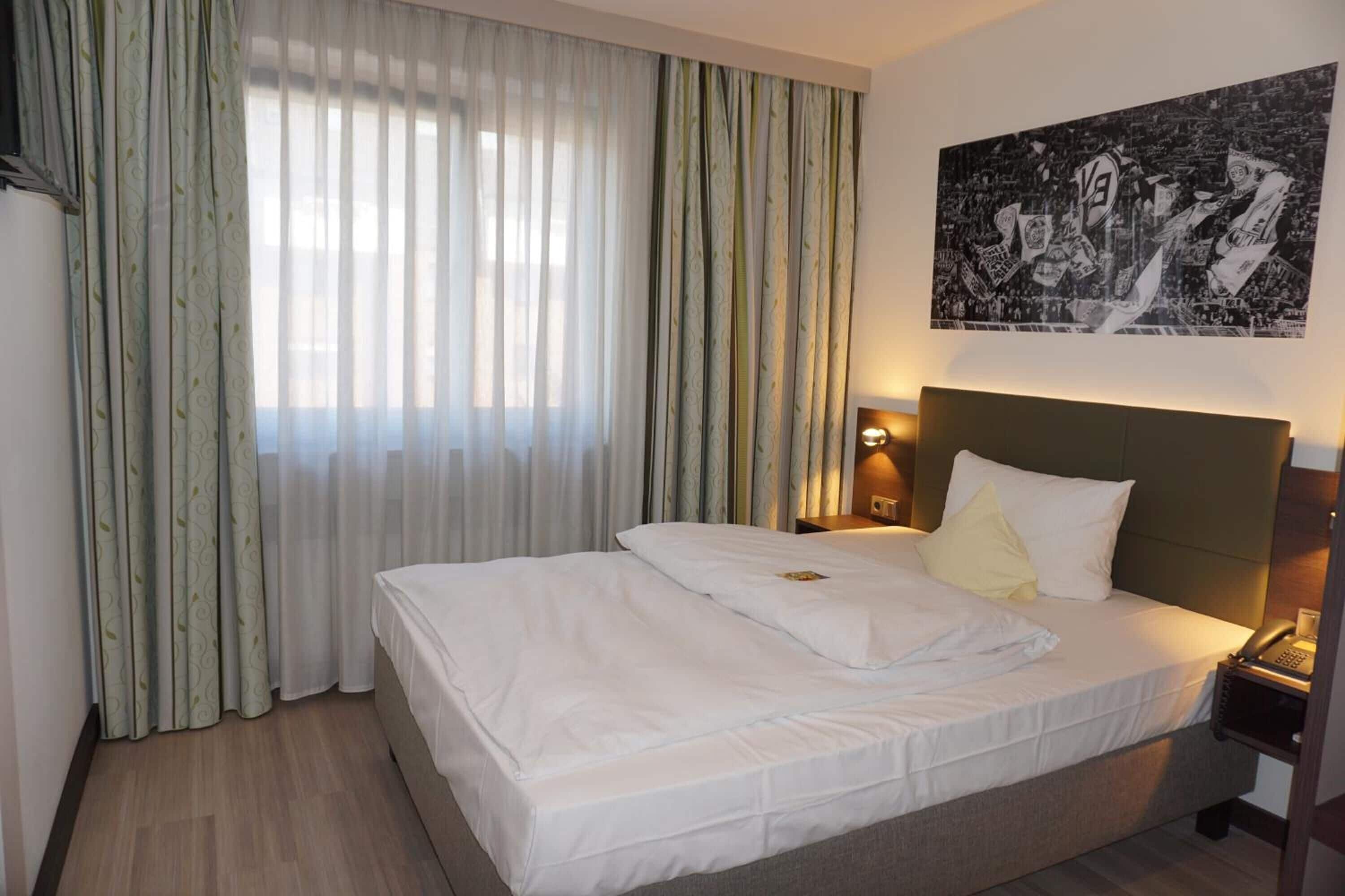 Quarto de hotel com uma cama, cortinas, luminária fixada na parede e um quadro emoldurado.
