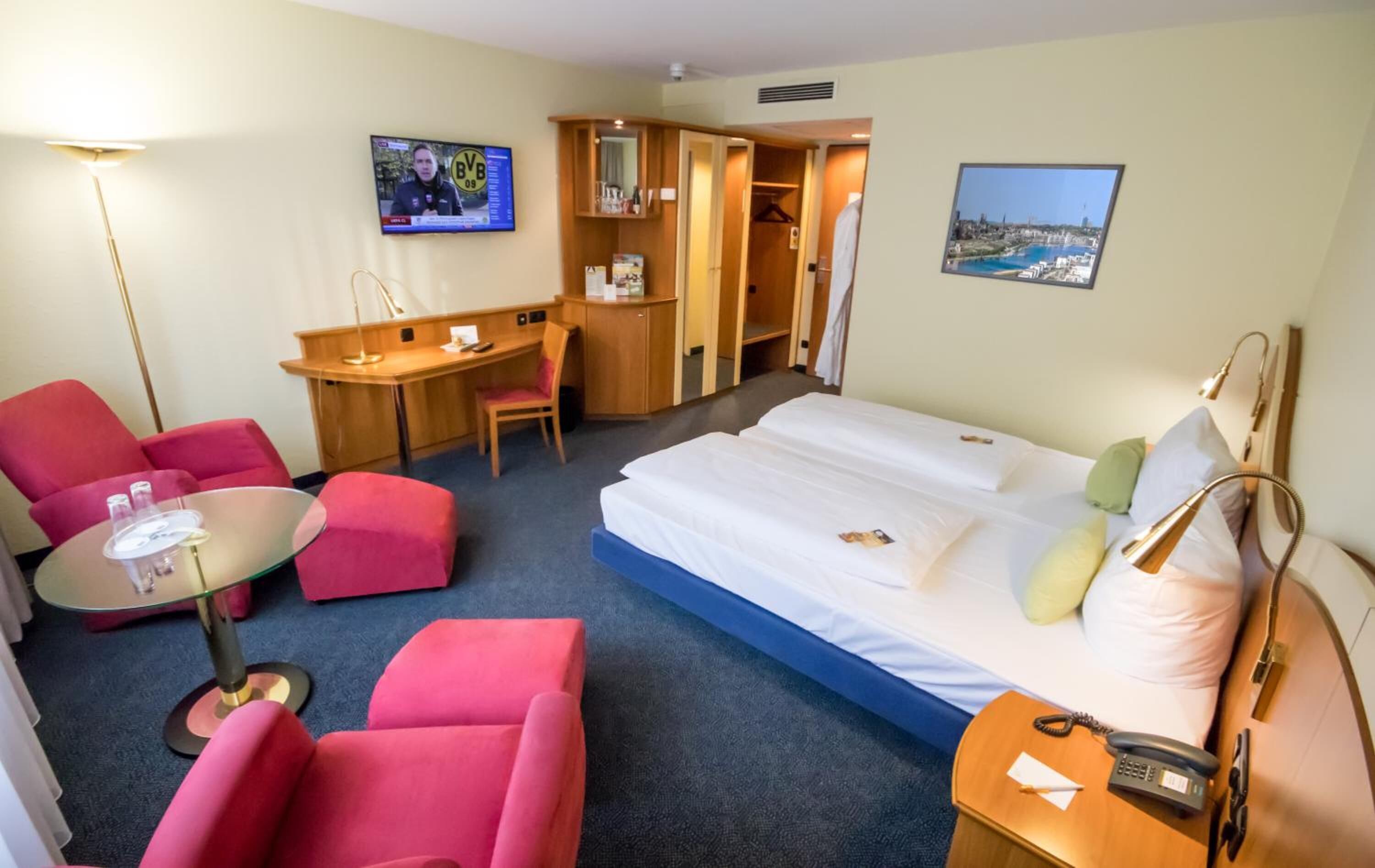 Quarto de hotel com cama, escrivaninha, cadeira, mesa pequena e televisão.