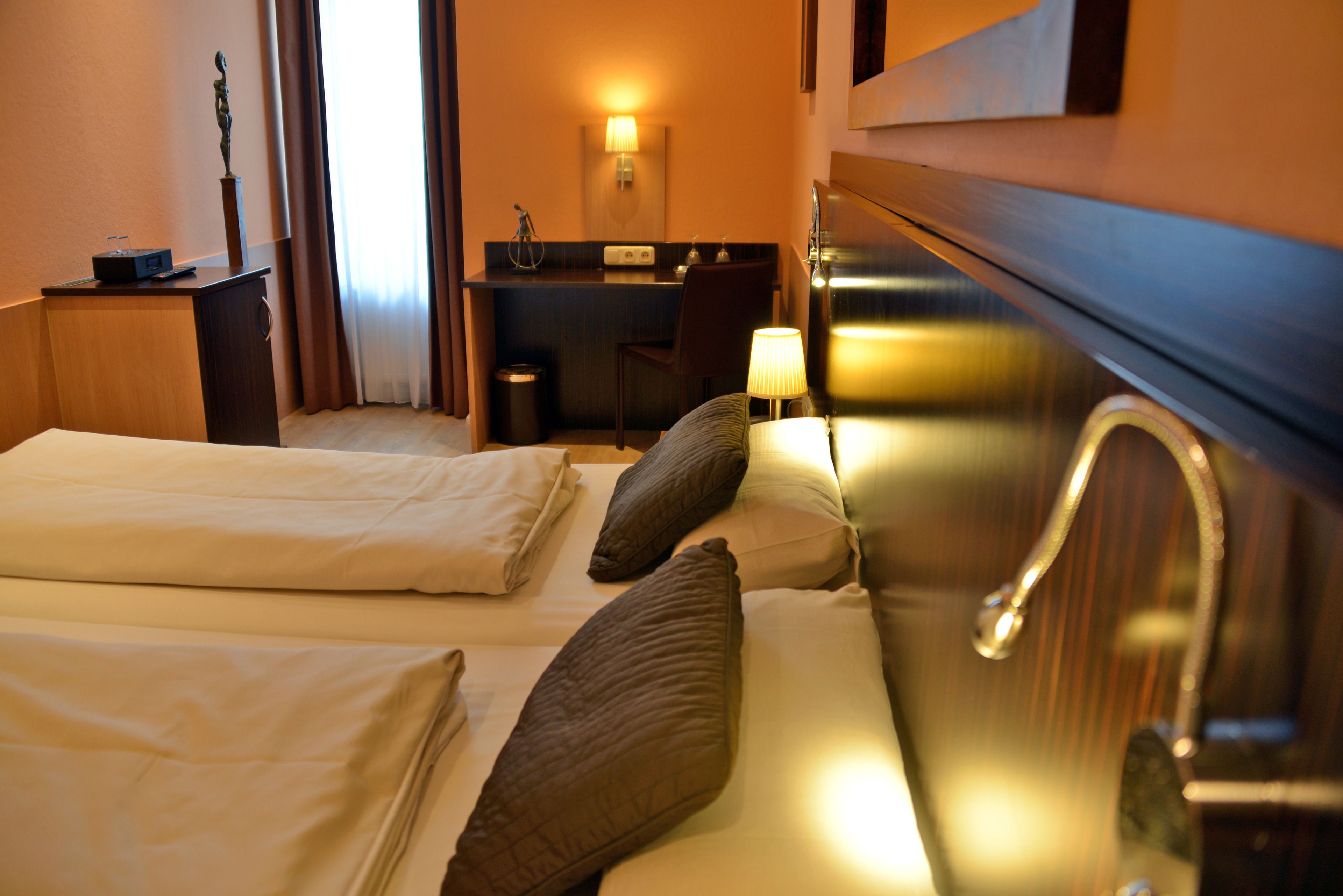 Quarto de hotel com duas camas, uma mesa, uma cadeira, um abajur e uma lixeira.