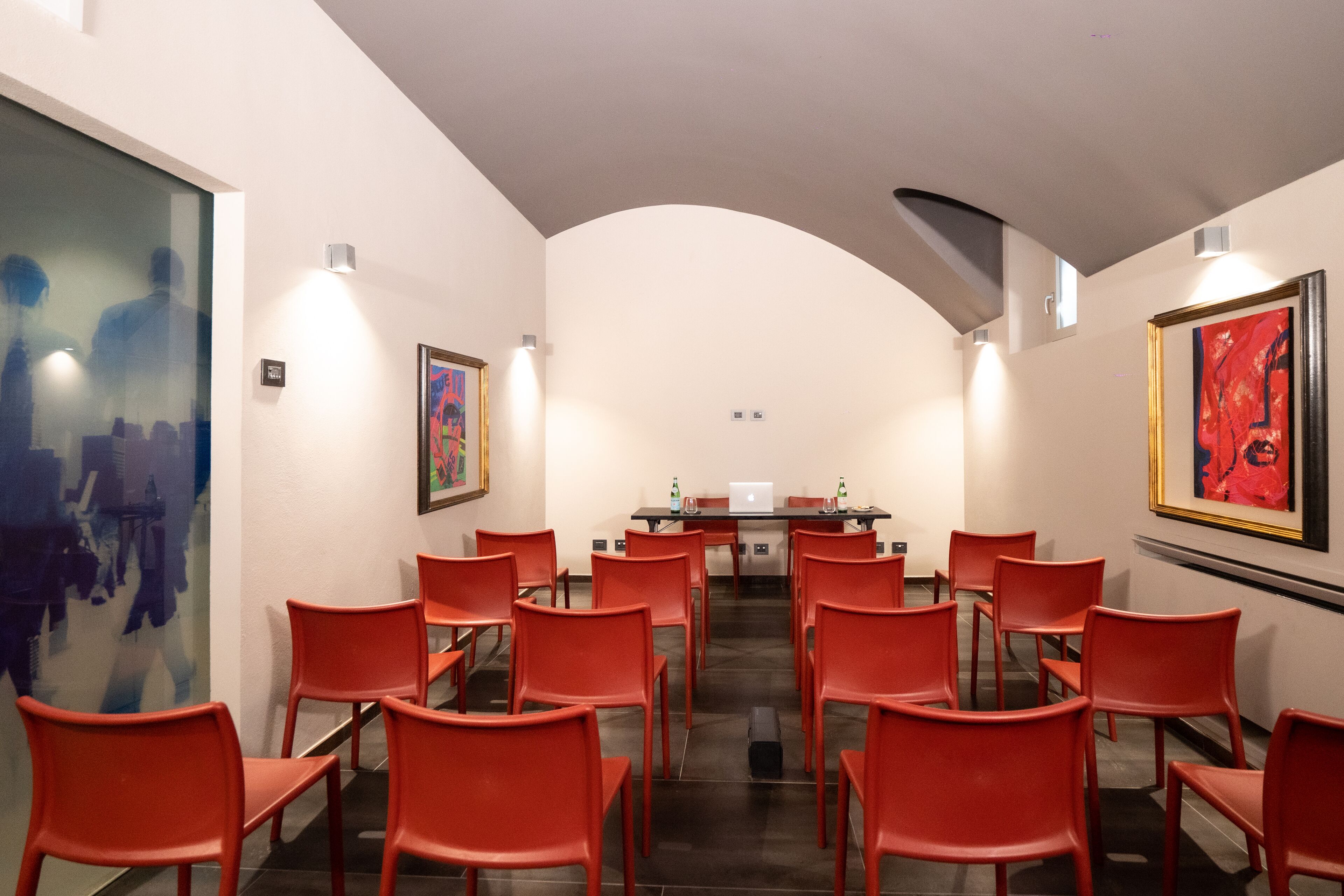 Uma sala de conferências com cadeiras vermelhas dispostas em fileiras, uma mesa com um laptop e quadros nas paredes.
