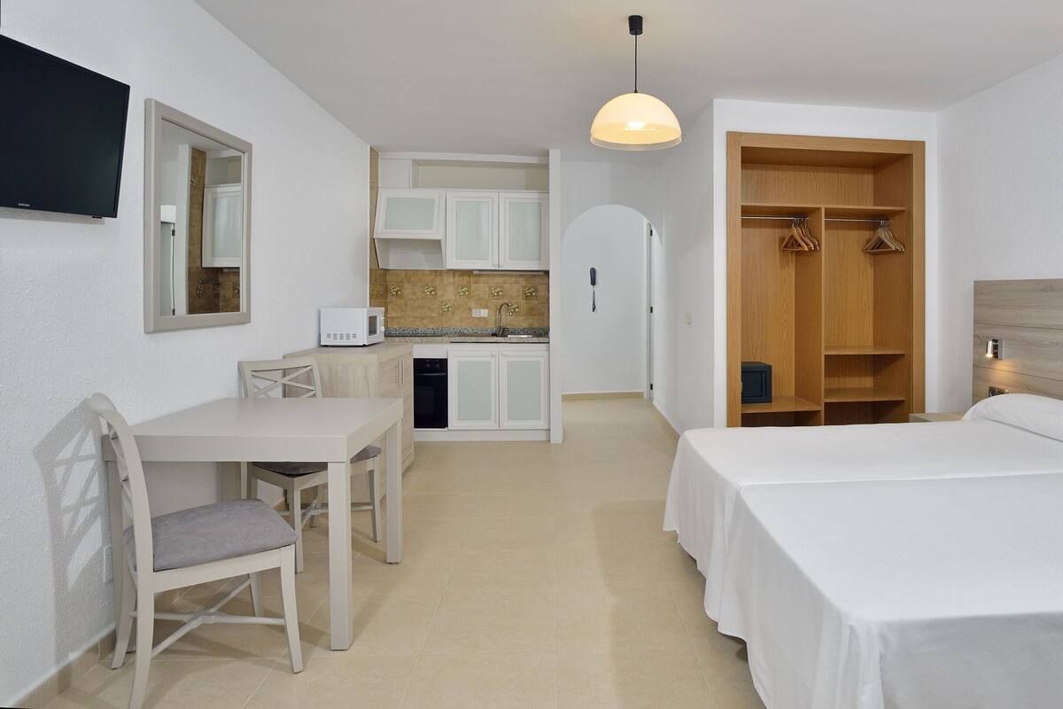 Quarto de hotel com cama, escrivaninha, cadeira, cozinha compacta e guarda-roupa.