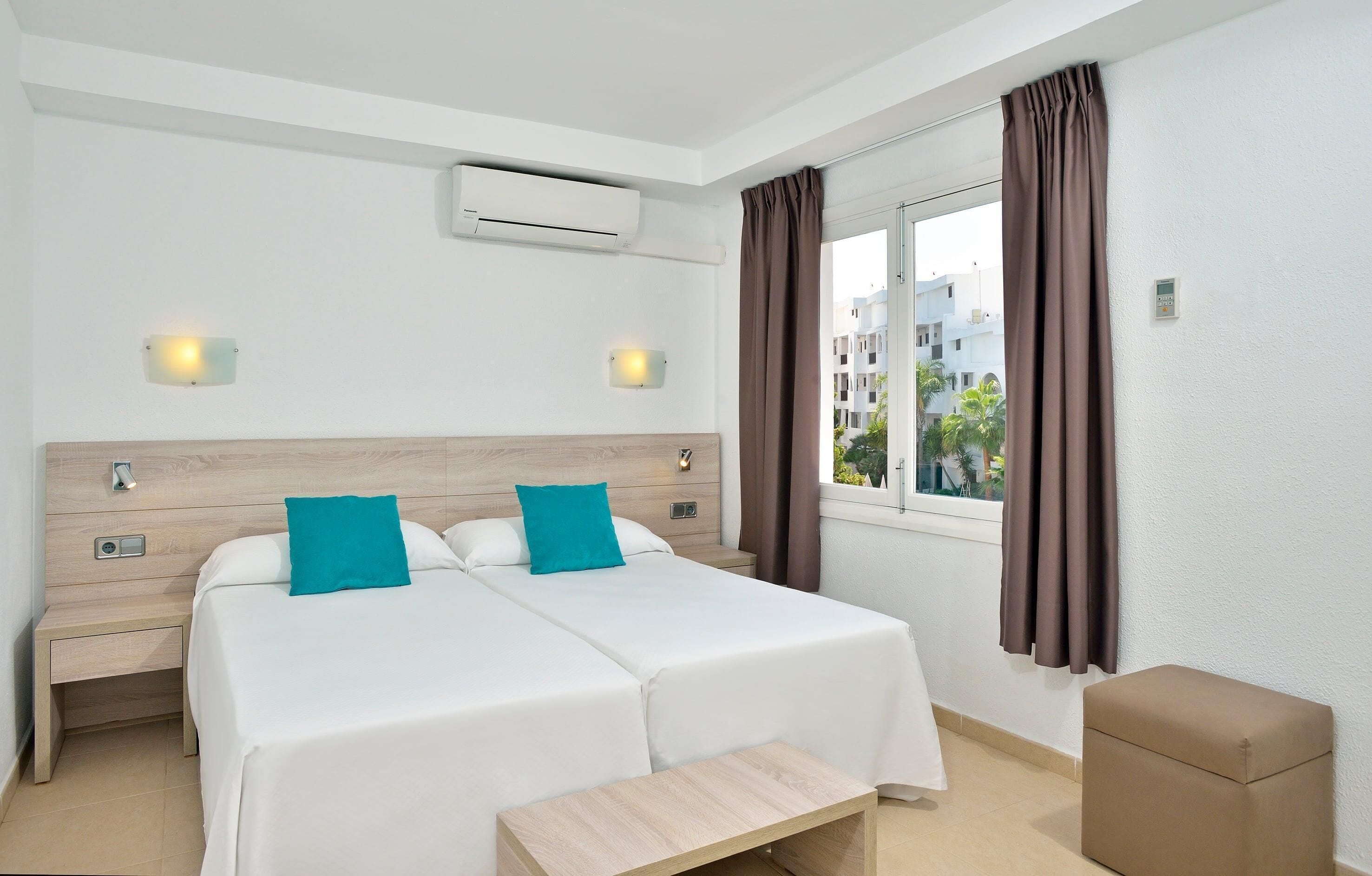 Quarto de hotel com duas camas, uma mesinha de cabeceira, um pequeno banco de madeira, um aparelho de ar condicionado e uma janela com cortinas.