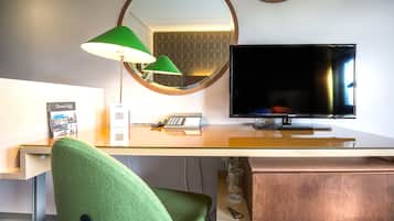 Quarto de hotel com escrivaninha de vidro, uma cadeira verde, televisão de tela plana, abajur, telefone e espelho.