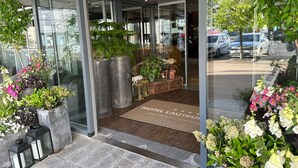 Entrada de hotel com porta de vidro, banco de madeira e plantas em vasos.