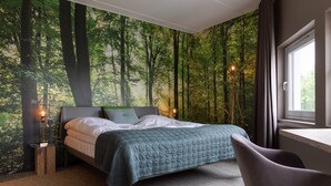 Quarto com papel de parede com tema de floresta, cama com manta verde, mesa de cabeceira com abajur e janela com cortinas.