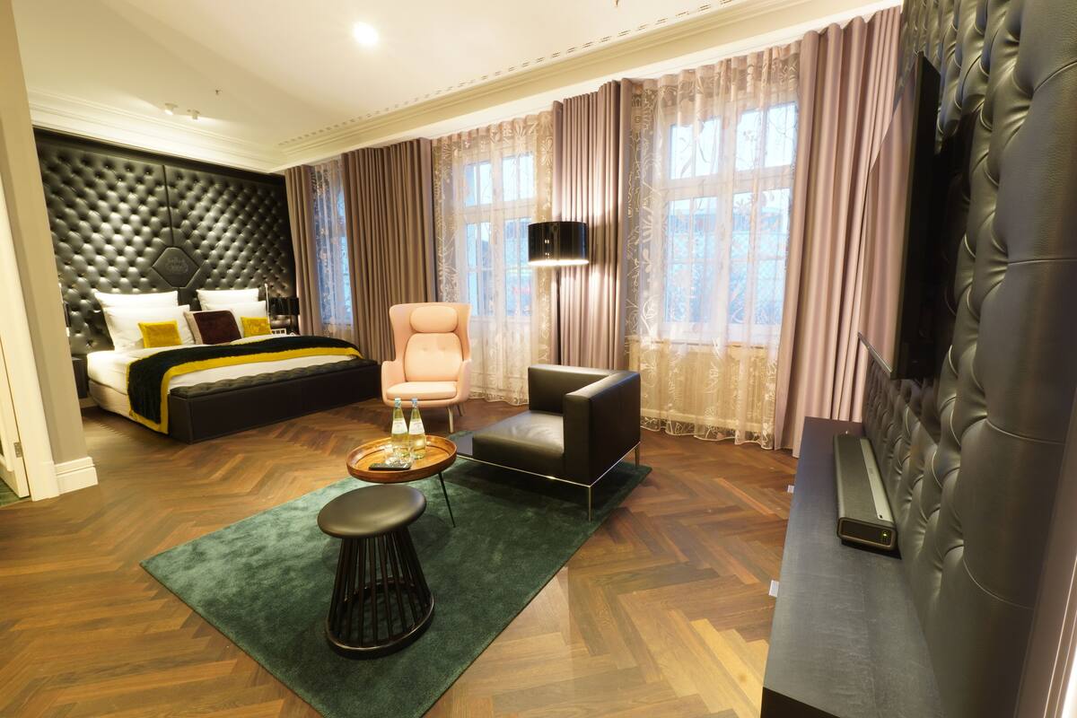 Quarto de hotel moderno com cabeceira acolchoada, uma cama grande com roupa de cama amarela e preta, uma poltrona rosa, um sofá preto, um tapete verde e uma televisão de tela plana.