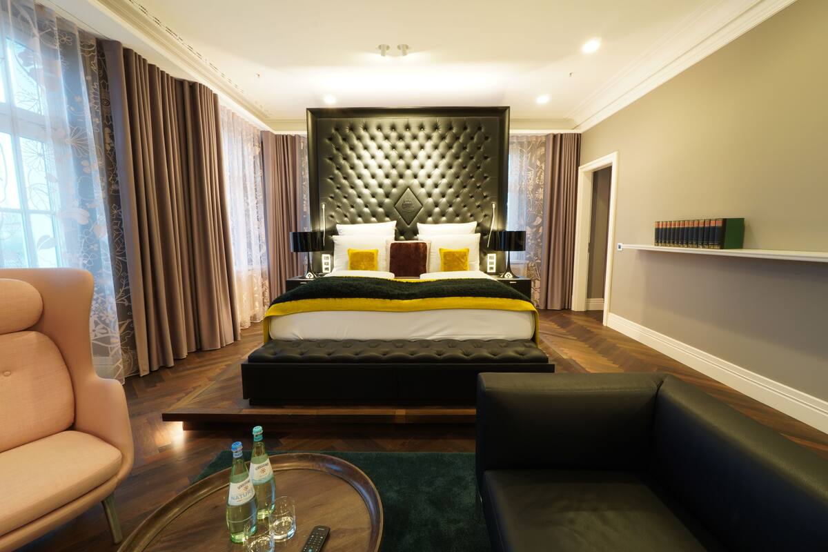 Quarto de hotel moderno com uma cama grande, um sofá, uma mesa de centro com garrafas e uma estante de livros.