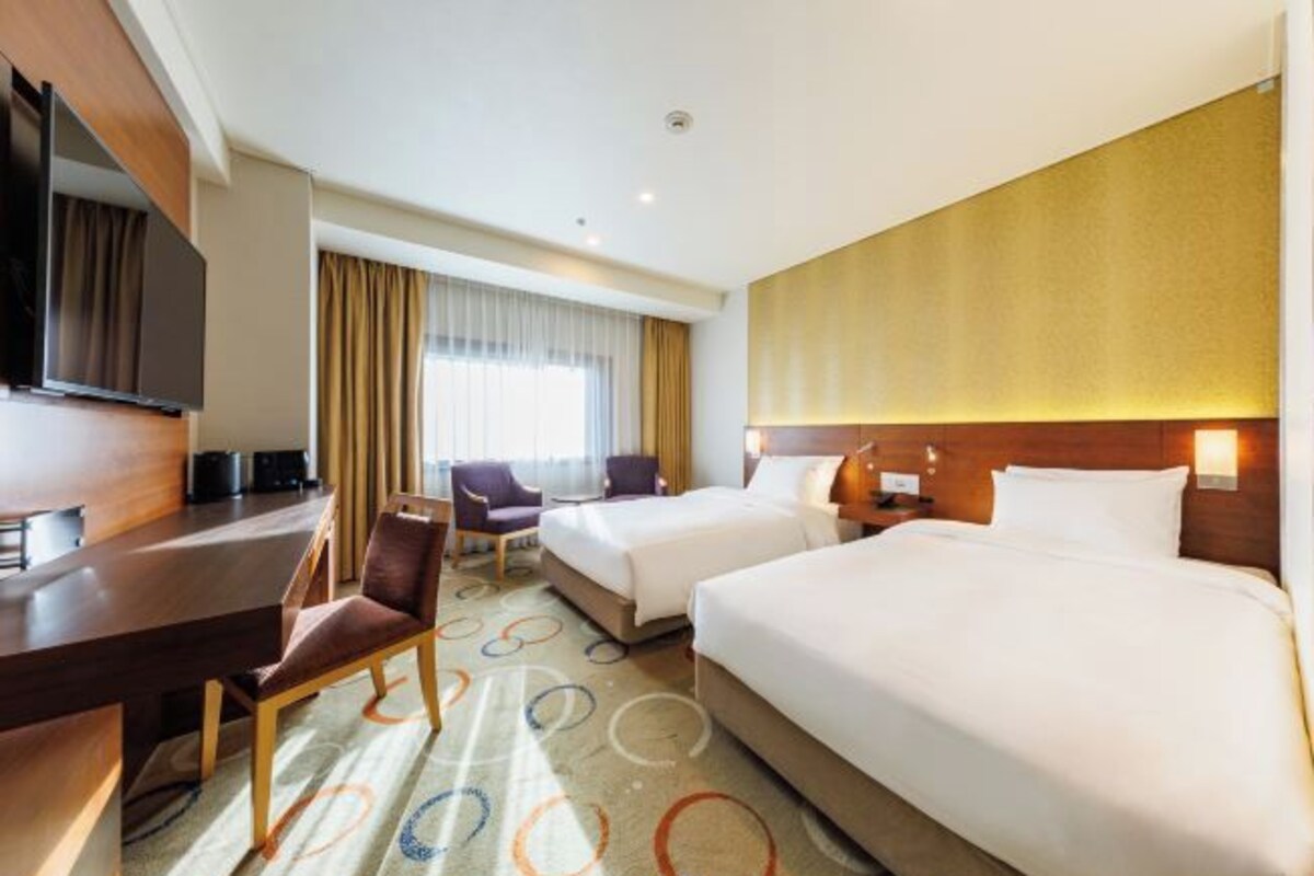 Quarto de hotel com duas camas, uma escrivaninha, uma cadeira, uma televisão e uma janela grande com cortinas.