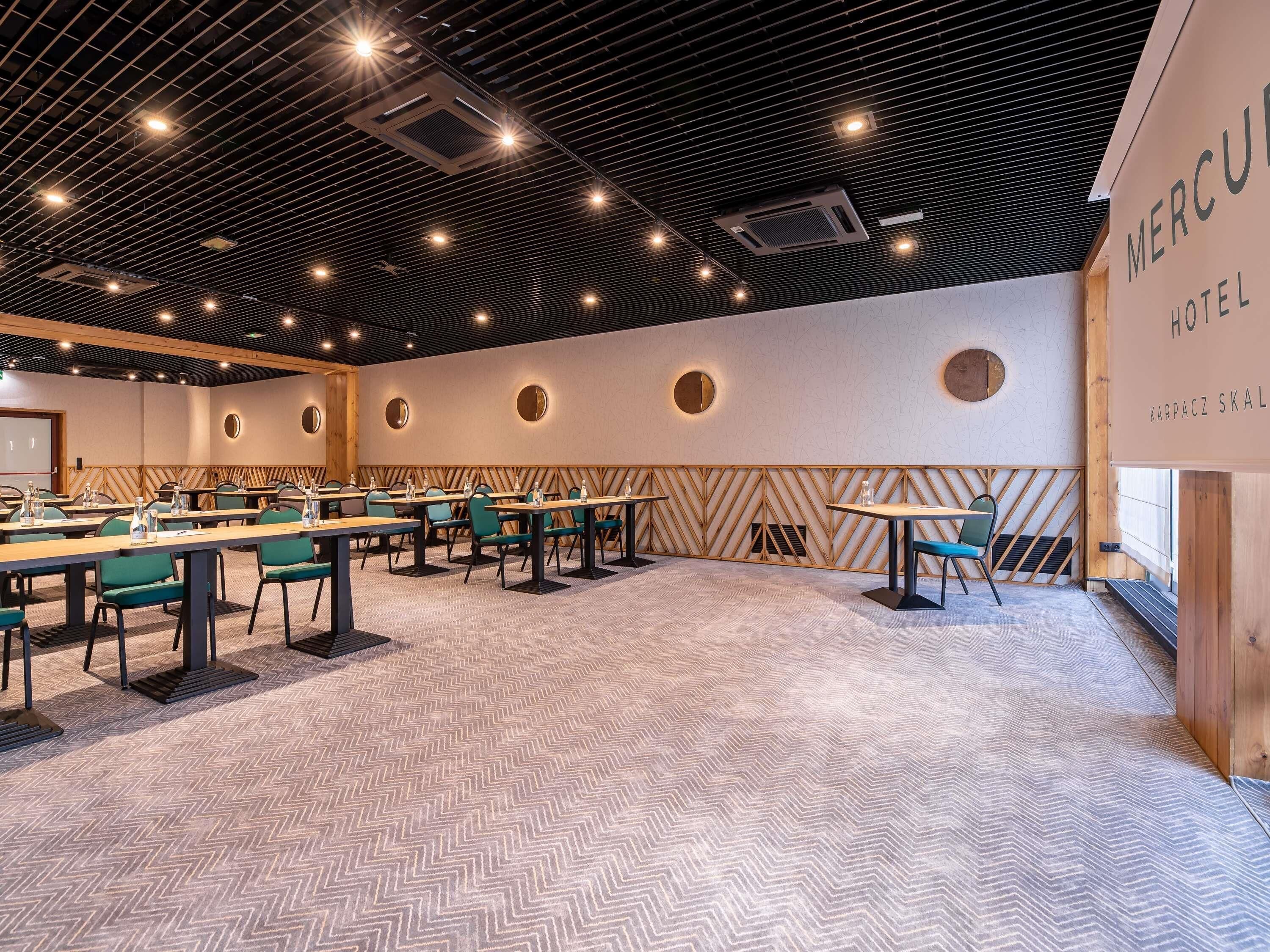 Uma sala de conferências com mesas, cadeiras, um painel de madeira e teto com iluminação embutida.