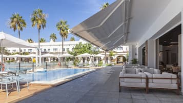 Uma área de piscina com espreguiçadeiras e guarda-sóis, palmeiras e um edifício com amplas janelas.