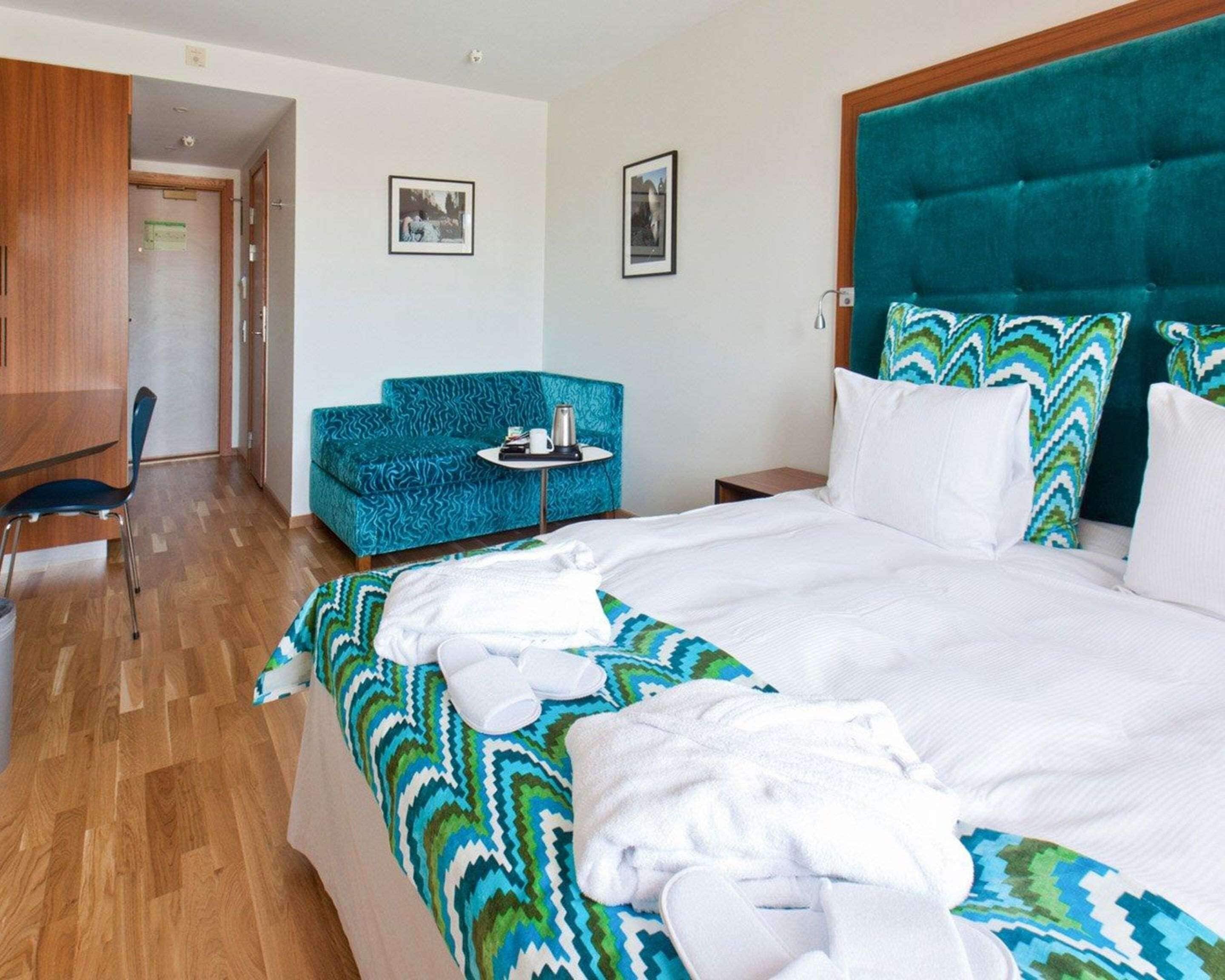 Quarto de hotel com uma cama grande, um sofá azul, uma pequena mesa com chaleira e uma cadeira.