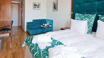 Quarto de hotel com uma cama grande, um sofá azul, uma pequena mesa com chaleira e uma cadeira.
