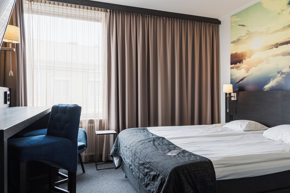 Quarto de hotel com uma cama, uma escrivaninha, uma cadeira, um abajur e uma pintura de um pôr do sol sobre uma paisagem.
