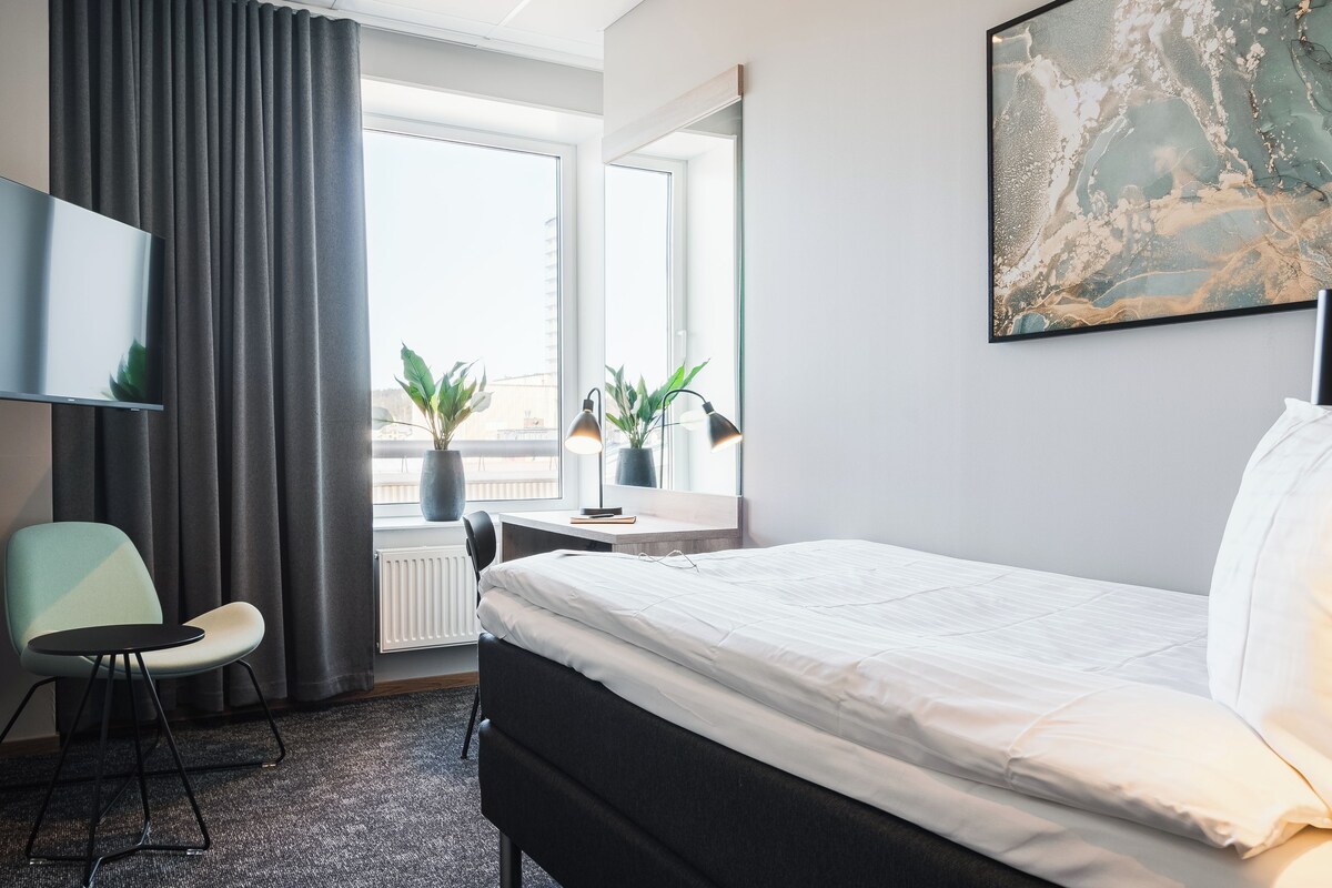 Quarto de hotel com cama, escrivaninha, cadeira, televisão e janela com cortinas.