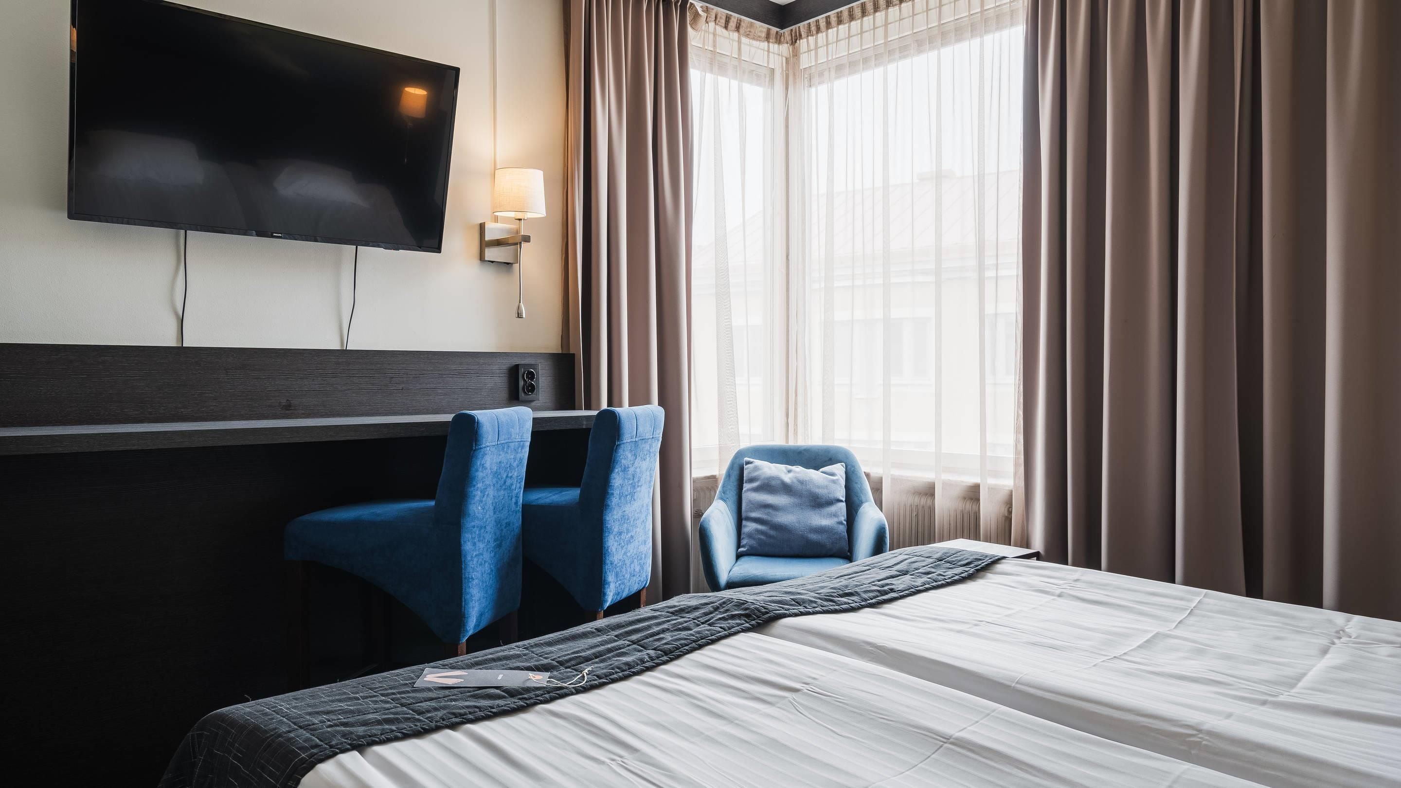 Quarto de hotel com cama, escrivaninha, cadeira, televisão e janela com cortinas.