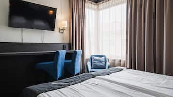 Quarto de hotel com cama, escrivaninha, cadeira, televisão e janela com cortinas.