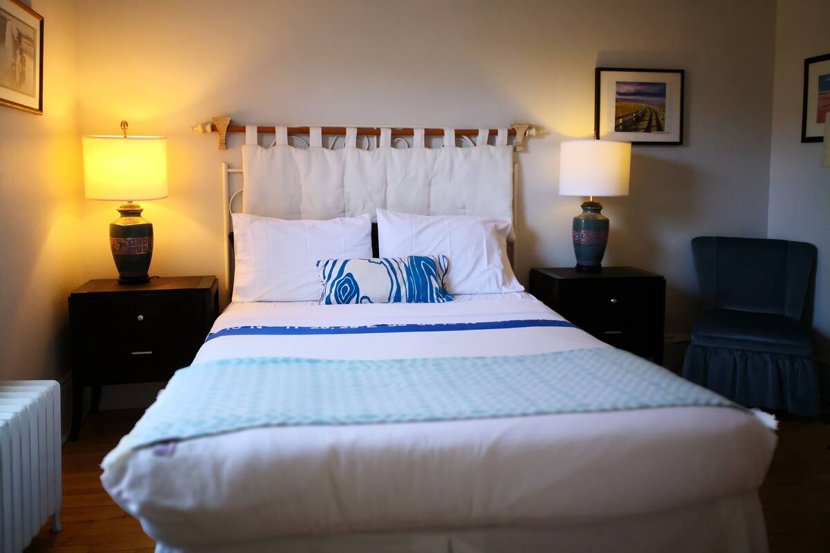 Cama bem arrumada com roupa de cama branca e detalhes azuis, acompanhada por duas mesas de cabeceira com luminárias, uma cadeira e quadros nas paredes.
