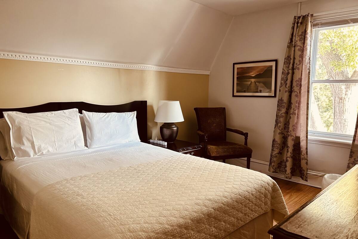 Cama bem arrumada com lençóis brancos, luminária de cabeceira, cadeira e quadro na parede.