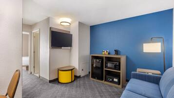 Quarto de hotel com parede azul de destaque, televisão de tela plana na parede, pequeno armário com dispositivos eletrônicos, sofá e poltrona.