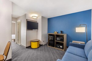 Quarto de hotel com parede azul de destaque, televisão de tela plana na parede, pequeno armário com dispositivos eletrônicos, sofá e poltrona.
