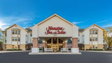Edifício do Hampton Inn & Suites com uma entrada ampla, pilares de tijolo e um céu azul claro.