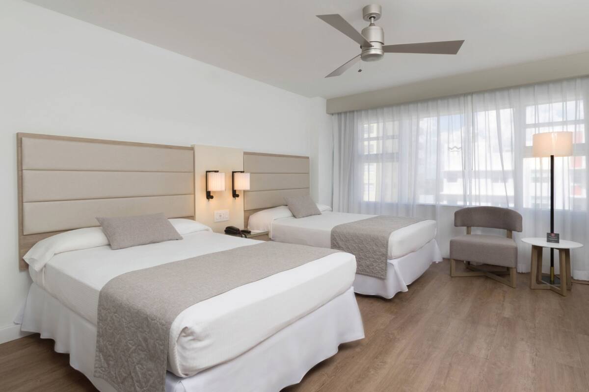 Quarto de hotel com duas camas, um ventilador de teto, mesa de cabeceira com luminária, uma cadeira e janela com cortinas finas.