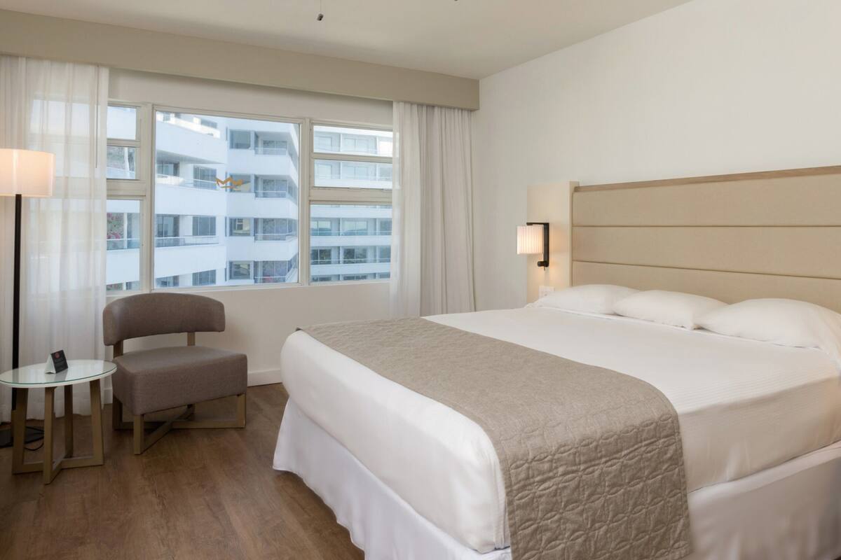 Quarto de hotel com uma cama grande, uma poltrona pequena, uma mesa de cabeceira e janela com vista para um prédio.