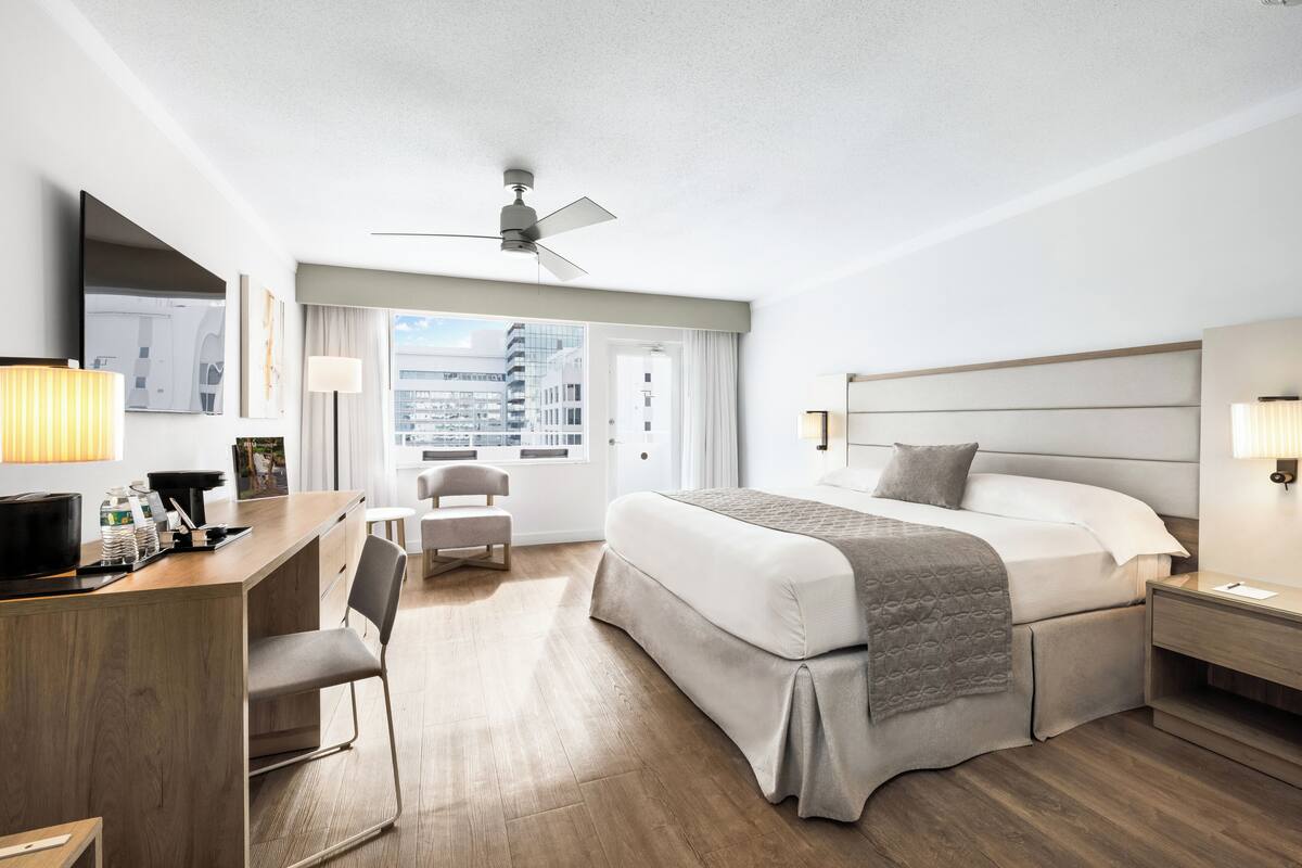 Quarto de hotel moderno com uma cama grande, uma escrivaninha, uma cadeira, um ventilador de teto e vista para a cidade pela janela.