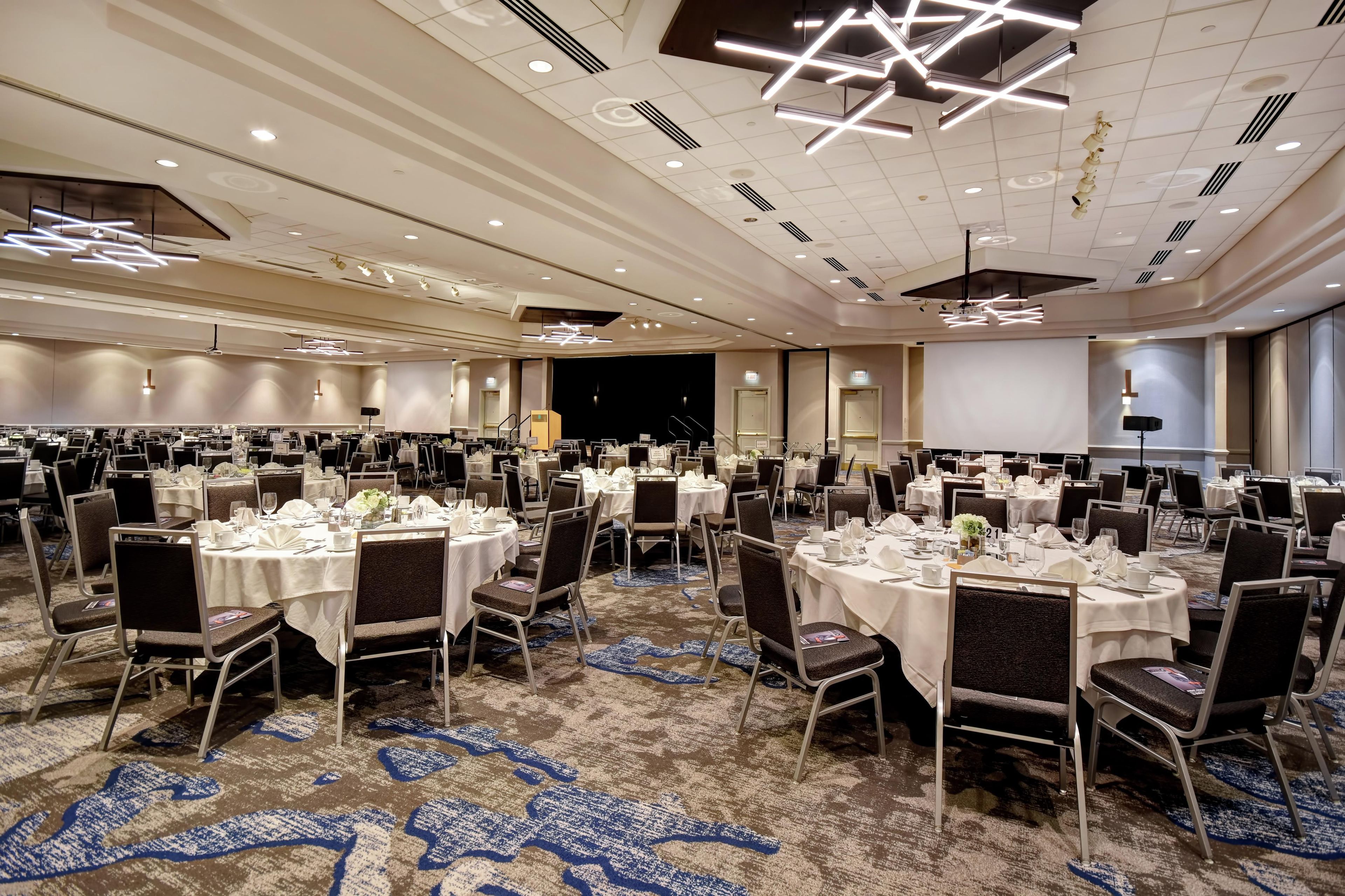 Uma grande sala de banquetes com mesas redondas dispostas para refeições, equipada com cadeiras e louças.