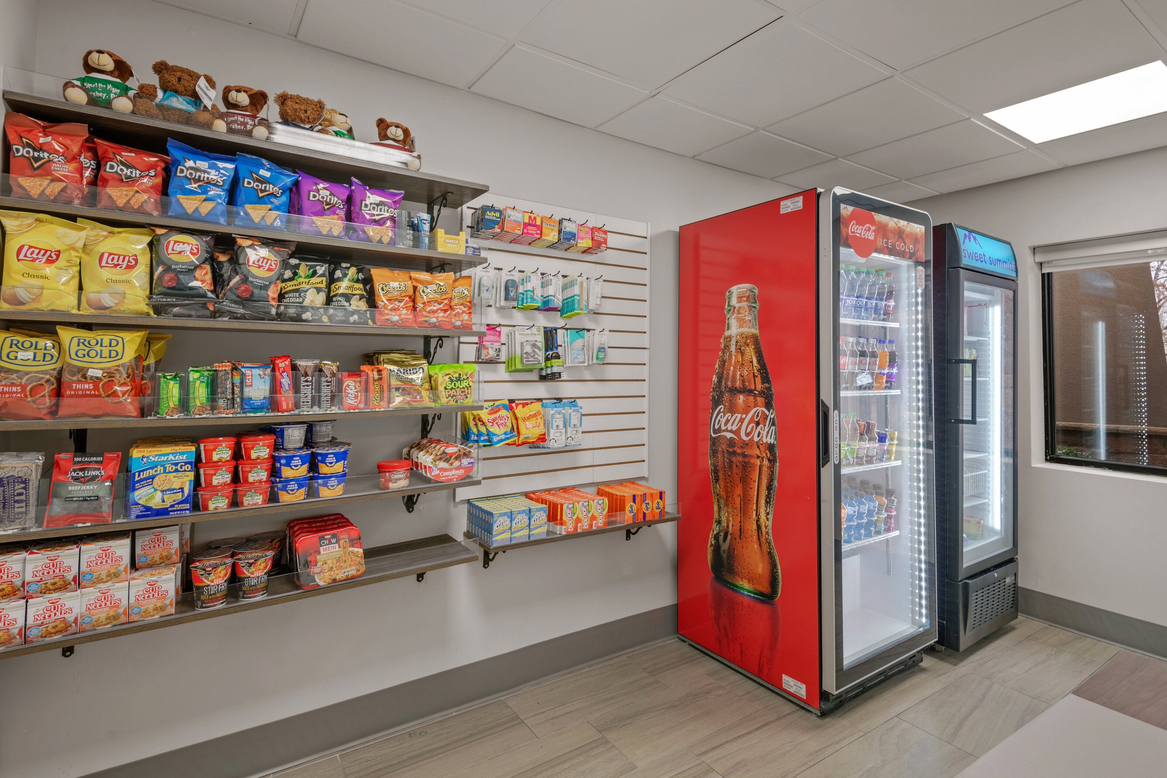Uma fileira de petiscos com diversas marcas de batatas e salgadinhos, um refrigerador de Coca-Cola e uma janela com persianas.