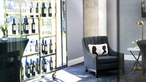 Um quarto com uma poltrona cinza escura, uma vitrine de vidro com garrafas de vinho, um pilar de mármore e uma pequena mesa redonda com um abajur.