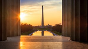 Vista do Monumento a Washington ao pôr do sol, emoldurado por colunas imponentes, com um lago refletor em primeiro plano.