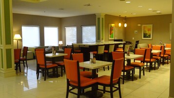 Espaço de refeições de hotel com várias mesas e cadeiras, um balcão de recepção e janelas amplas.