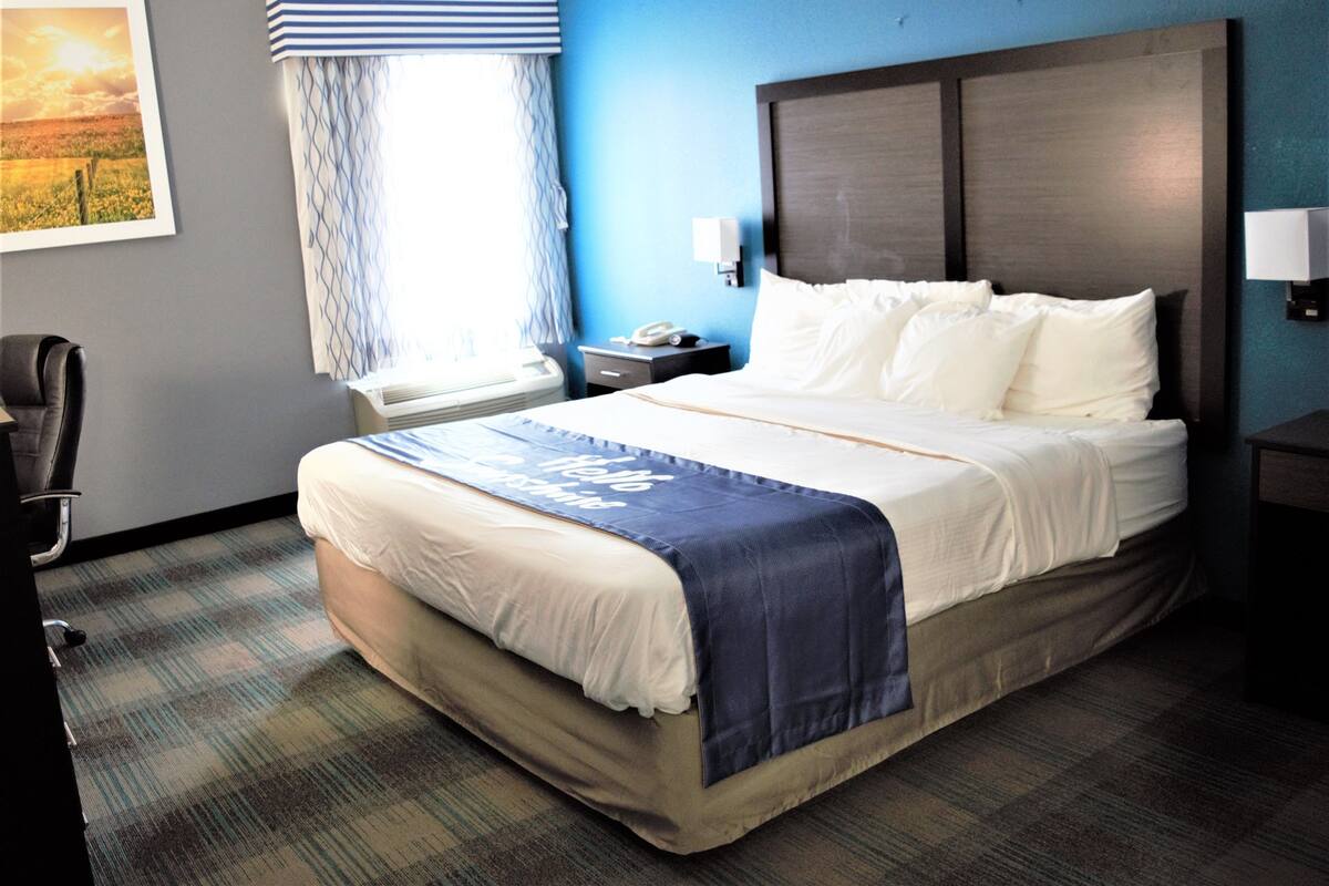 Quarto de hotel com cama, escrivaninha, cadeira, janela com cortinas e luminária na parede.