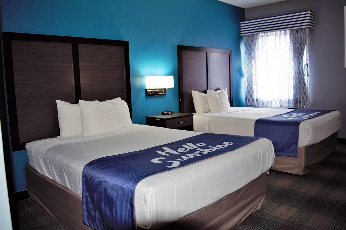 Quarto de hotel com duas camas, cada uma com uma colcha estampada com a inscrição 'Hello Sunshine', uma mesa de cabeceira com abajur e uma janela com cortinas.