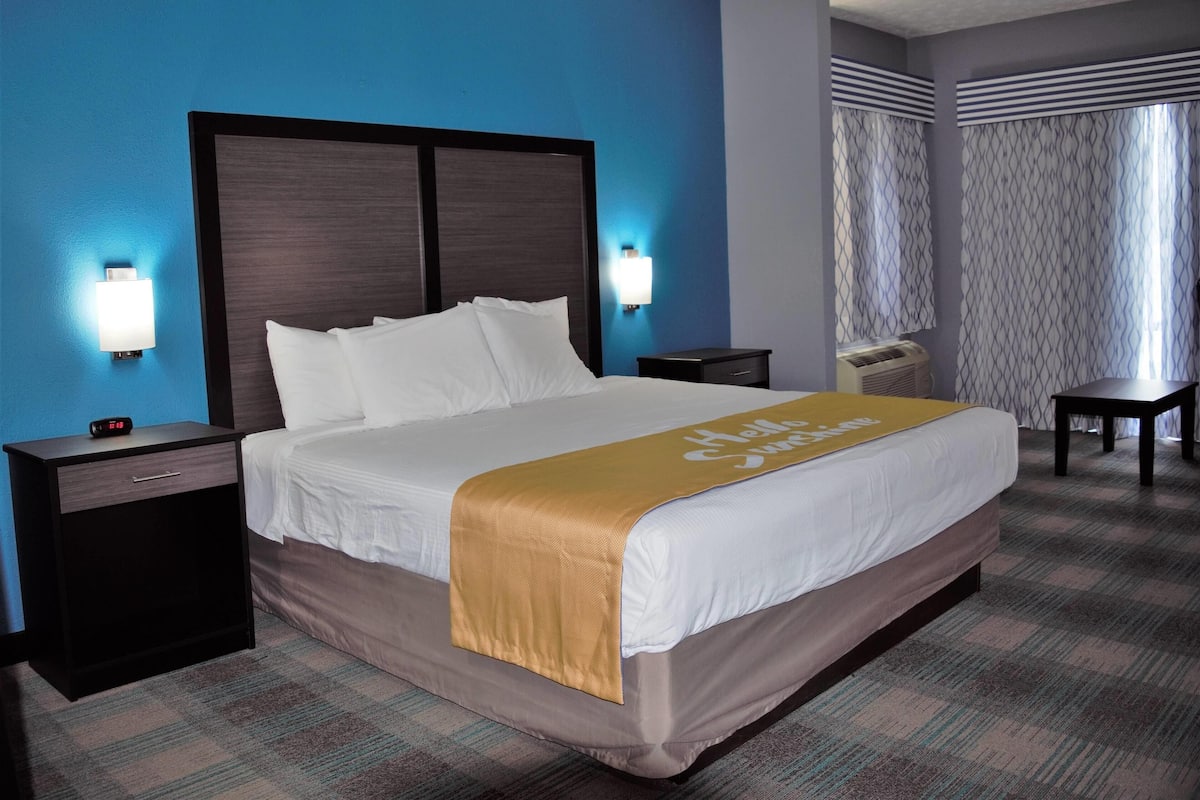 Quarto de hotel com uma cama grande, duas mesas de cabeceira, um criado-mudo, um relógio de parede, um termostato e uma janela com cortinas.