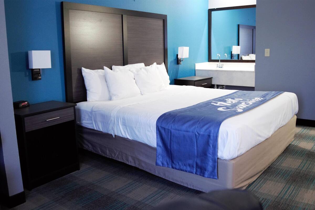 Quarto de hotel com uma cama grande, mesa de cabeceira, cômoda, banheiro com espelho e parede azul.
