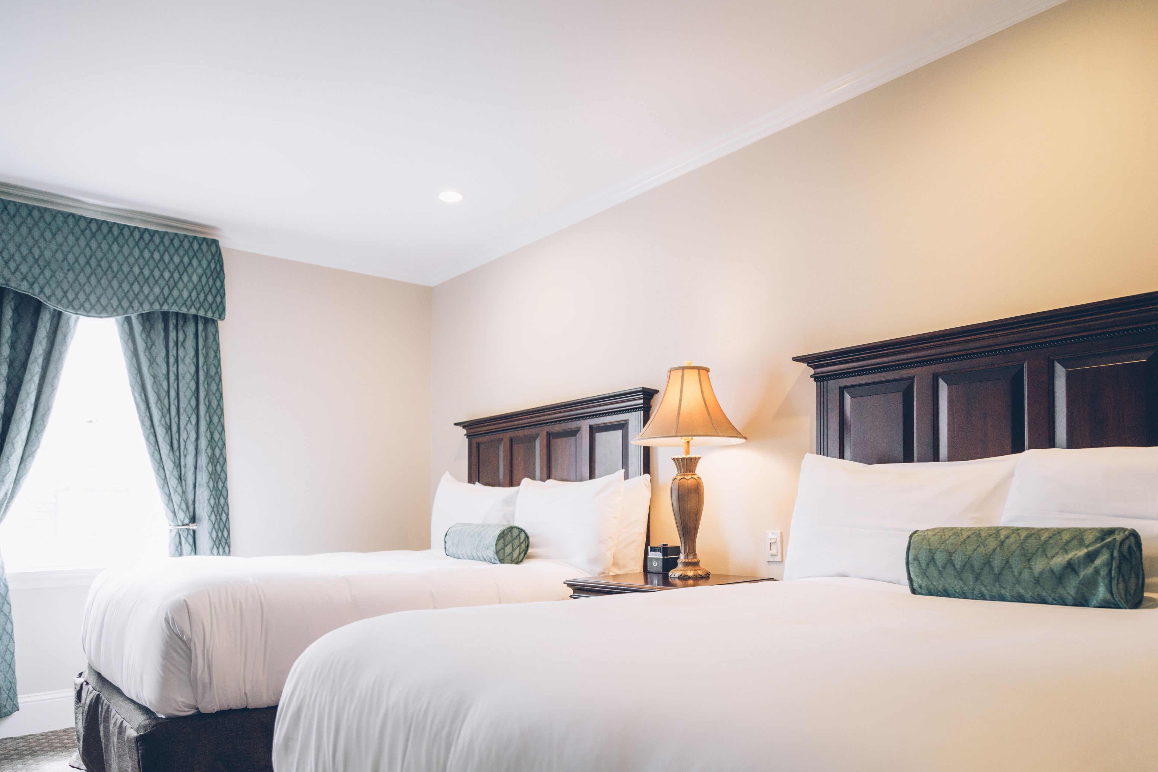 Quarto de hotel com duas camas, uma mesa de cabeceira com abajur e uma janela com cortinas.