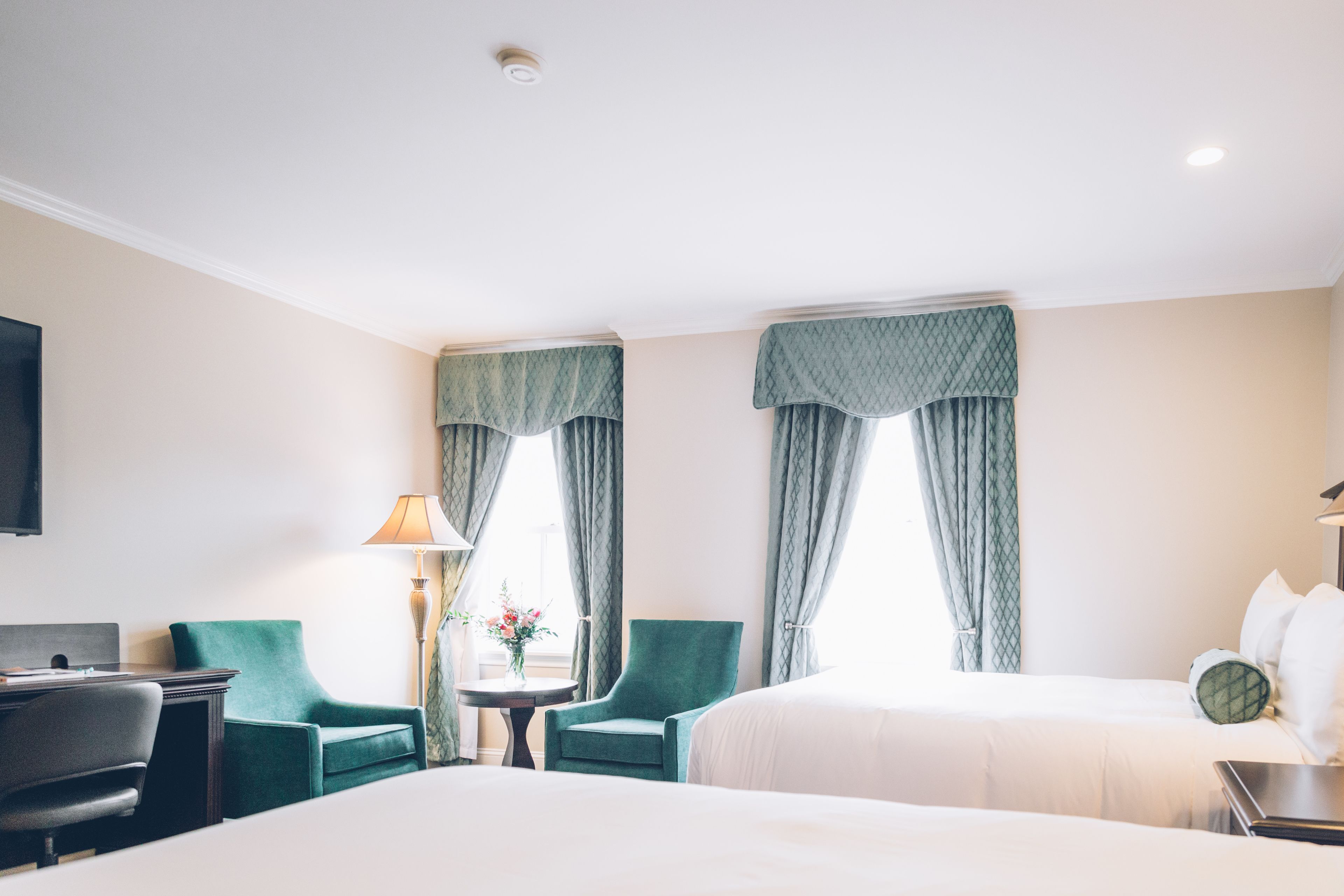 Quarto de hotel com uma cama, duas poltronas verdes, uma mesinha de centro com uma luminária e uma televisão na parede à esquerda.