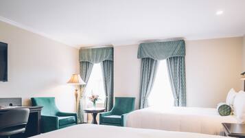 Quarto de hotel com uma cama, duas poltronas verdes, uma mesinha de centro com uma luminária e uma televisão na parede à esquerda.