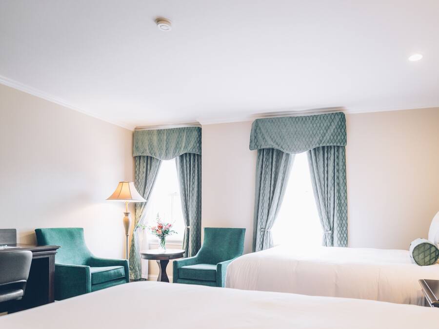 Quarto de hotel com uma cama, duas poltronas verdes, uma mesinha de centro com uma luminária e uma televisão na parede à esquerda.