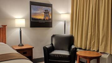 Quarto de hotel com uma cadeira de couro preto, duas mesas de cabeceira com luminárias, uma moldura com uma foto de um farol e uma janela com cortinas.