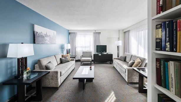 Quarto de hotel moderno com parede azul de destaque, um conjunto de sofás, uma TV de tela plana, uma mesa de centro e uma estante cheia de livros.